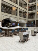 Um hall de hotel com vários níveis, uma área de bar central, espaços de estar e uma tela digital exibindo informações.