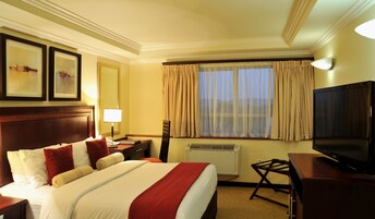 Quarto de hotel com cama, televisão, escrivaninha, luminária e janela com cortinas.