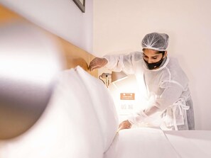 Uma pessoa vestida com bata branca e touca de cabelo, cuidando de uma cama em um quarto com uma placa na parede.