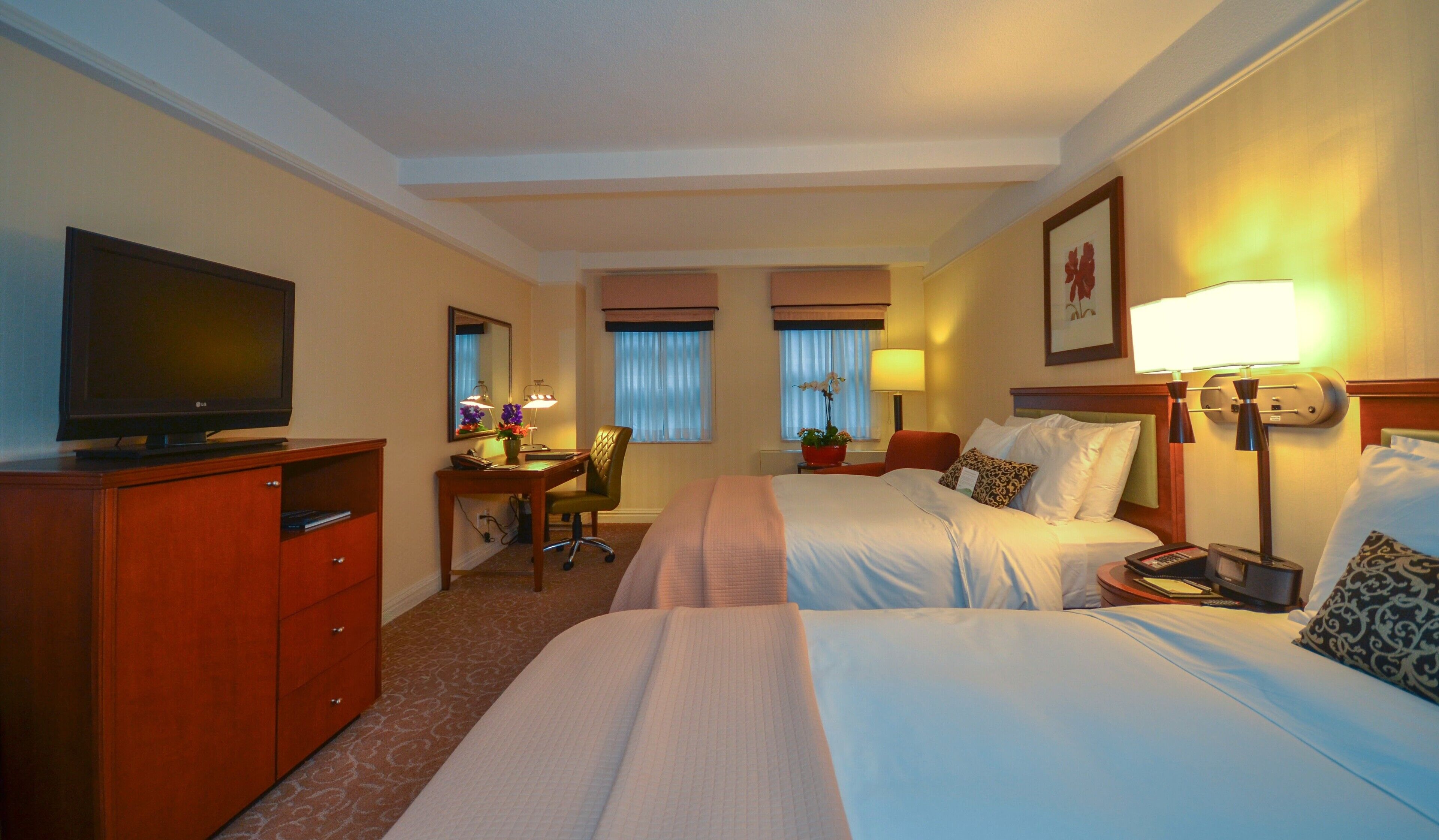 Quarto de hotel com duas camas, uma televisão, uma escrivaninha com cadeira e uma luminária.