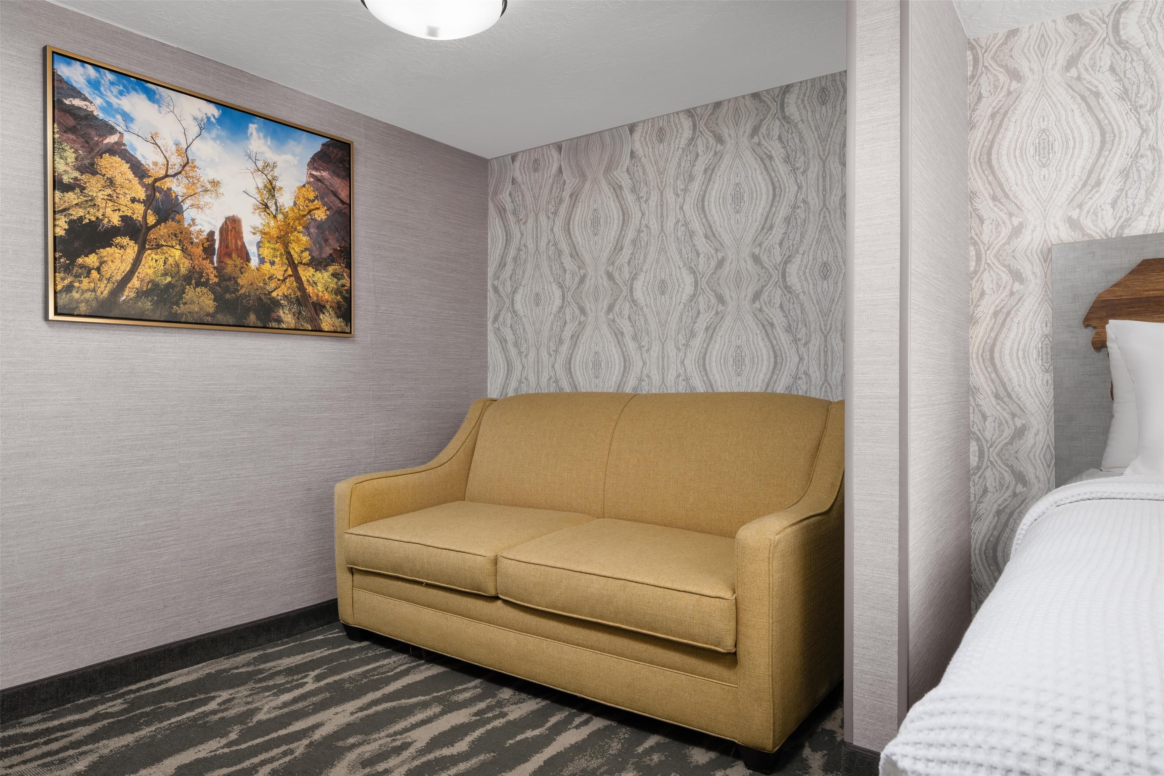 Quarto de hotel com uma grande moldura com uma imagem de um cânion, um sofá cor de mostarda, papel de parede estampado e uma cama com edredom branco.