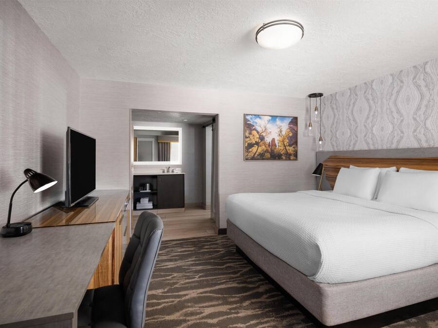 Quarto de hotel com cama, escrivaninha com televisão, luminária de mesa, cadeira, banheiro e um quadro na parede.
