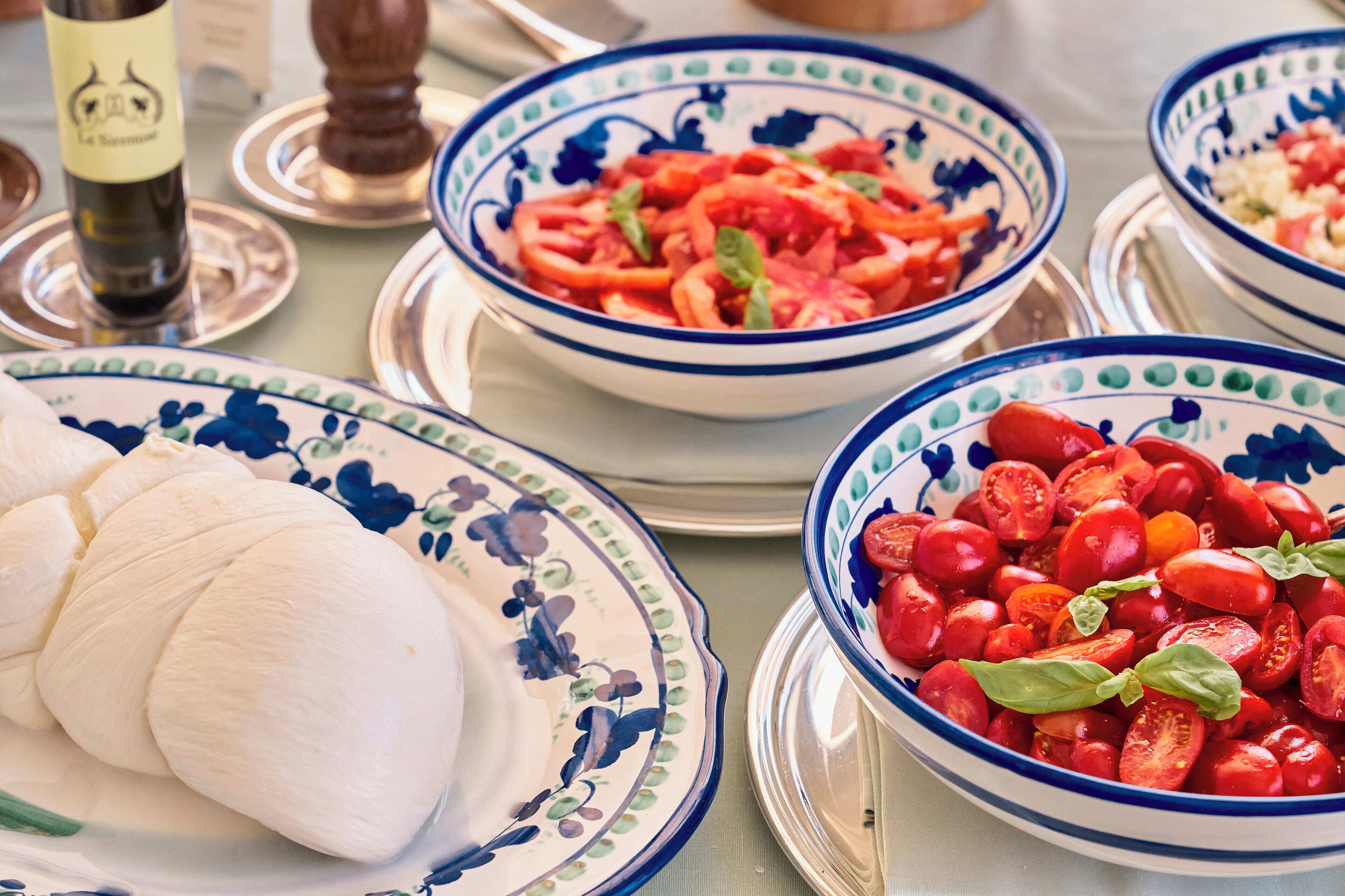 Uma mesa com diversos pratos, incluindo uma tigela de tomates fatiados com manjericão, uma porção de massa e uma garrafa de vinho.