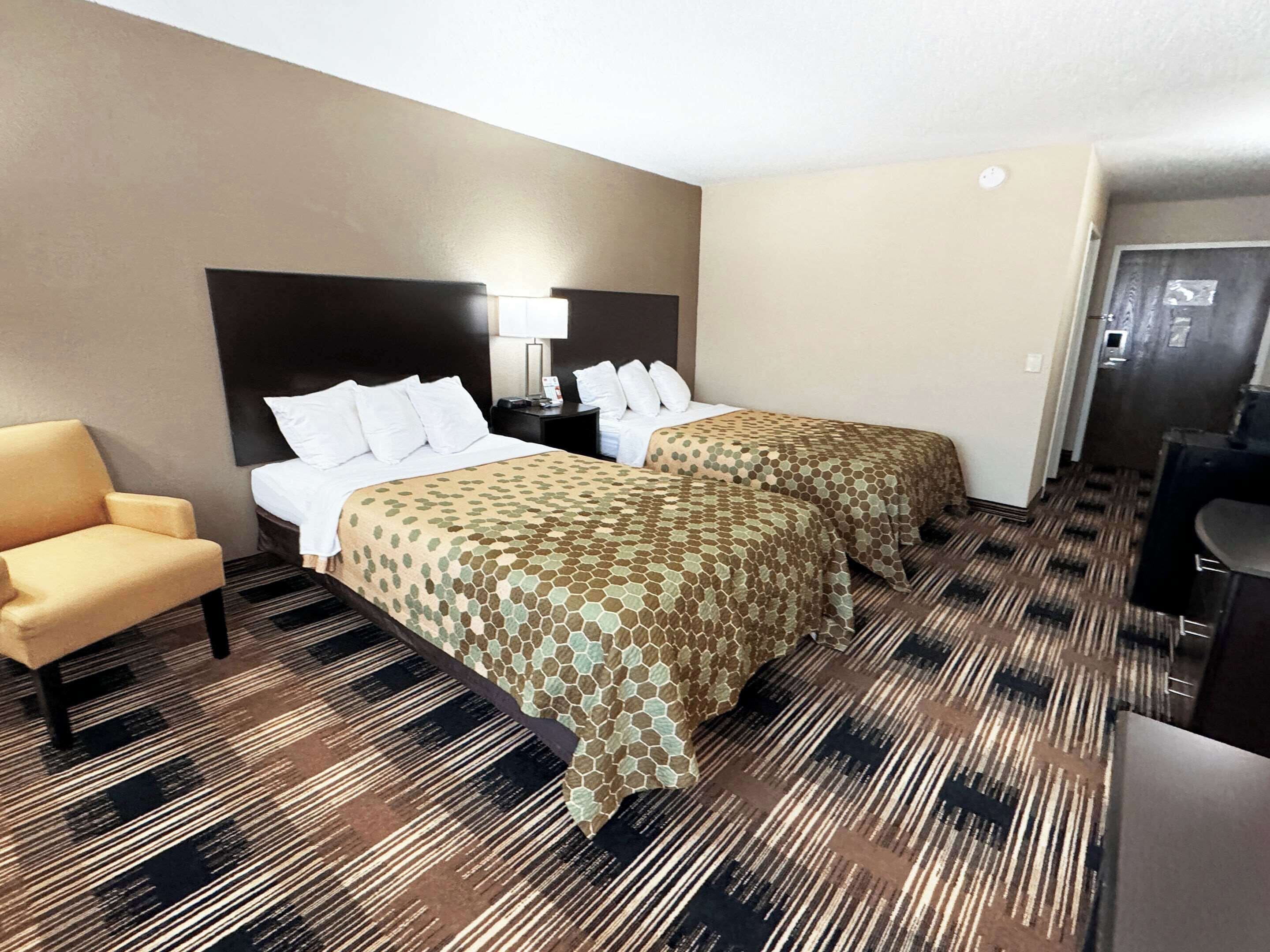 Quarto de hotel com duas camas, uma cadeira, uma escrivaninha e um abajur.
