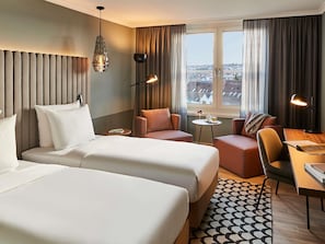 Quarto de hotel com duas camas, uma escrivaninha, uma cadeira, um abajur e vista para a cidade.
