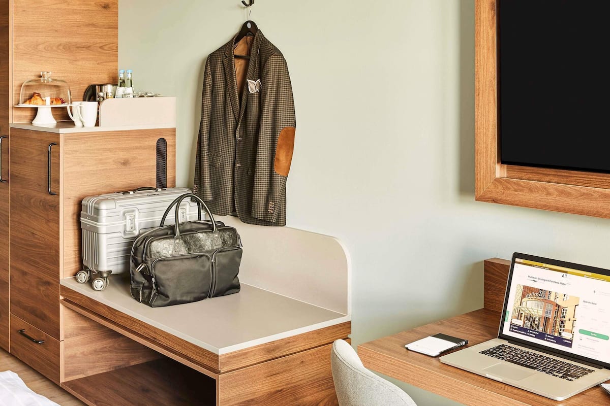Quarto de hotel com escrivaninha, um laptop, mala, bolsa, casaco pendurado e minibar.