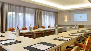 Sala de conferência com uma mesa comprida, cadeiras e tela de projeção exibindo 'Hotel Der Fohrenhof'.