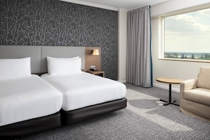 Quarto de hotel com duas camas, mesa de cabeceira, abajur, pequena mesa, sofá e uma grande janela com vista.