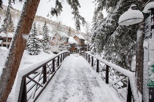 Uma ponte de madeira coberta por neve que leva a um edifício com telhado inclinado, cercado por árvores carregadas de neve.