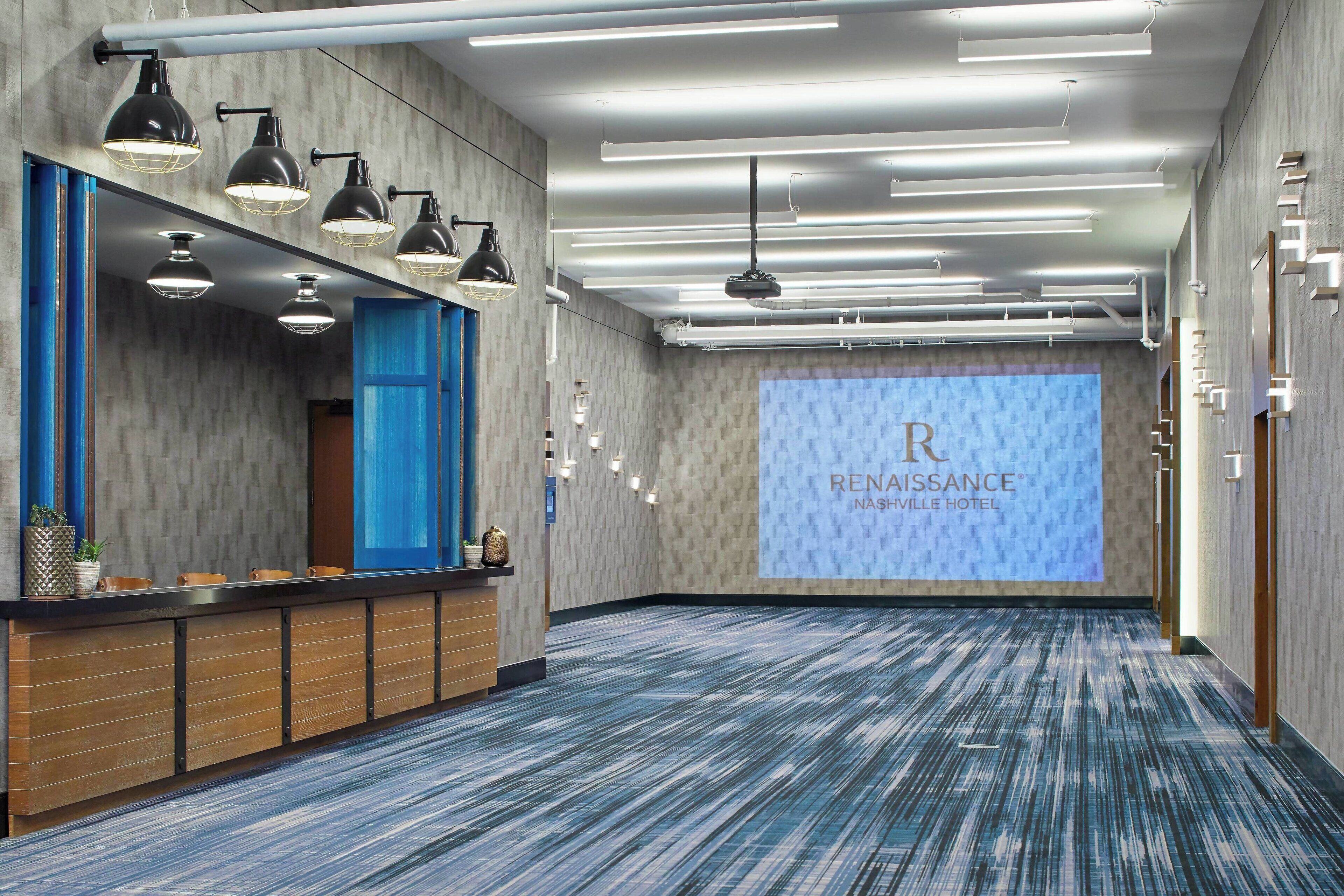 Uma área de recepção de hotel com design moderno, apresentando um balcão de atendimento, várias luminárias pendentes e uma tela grande exibindo o nome do hotel.