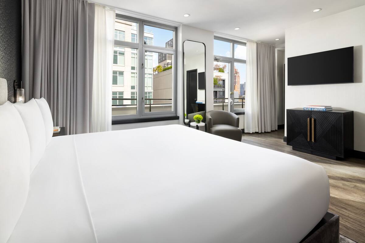 Quarto de hotel moderno com uma cama grande, uma cadeira, uma televisão e vista para a cidade.