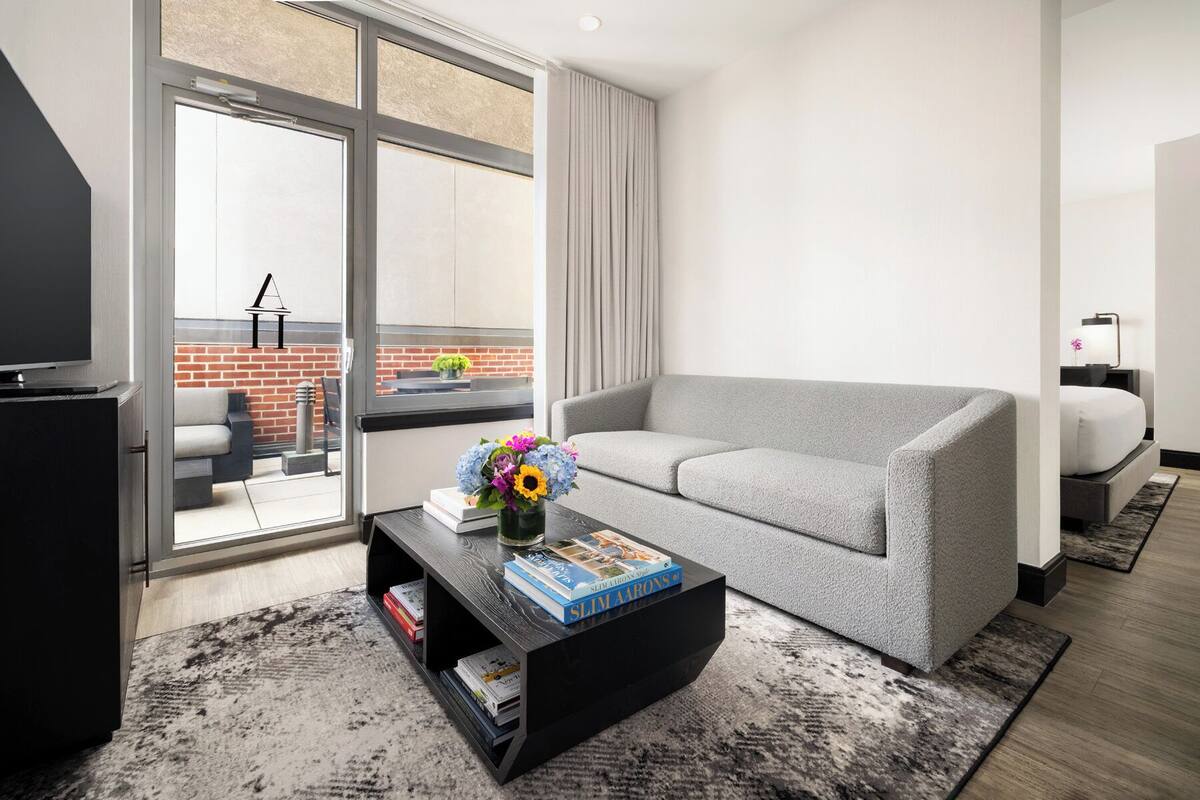 Uma sala de estar moderna com um sofá cinzento, uma mesa de centro com livros e um vaso de flores, uma TV sobre um móvel, e vista para um edifício de tijolos através de grandes janelas.