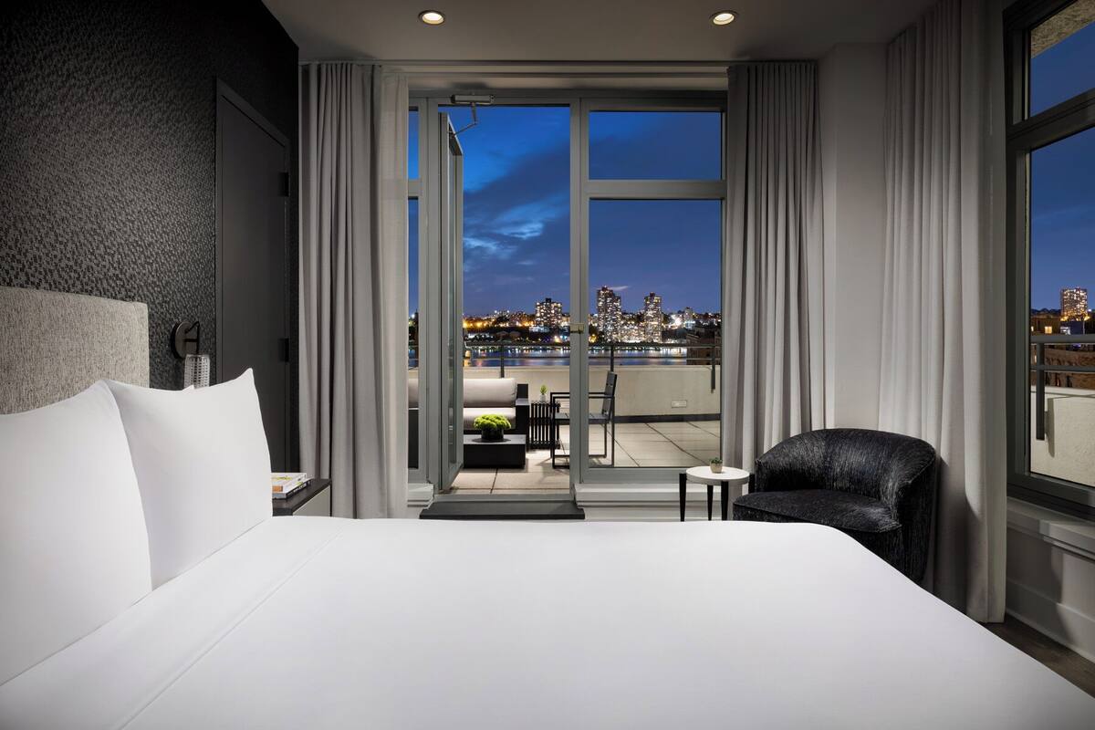 Quarto de hotel moderno com uma cama grande, varanda com assentos e vista para a cidade ao anoitecer.