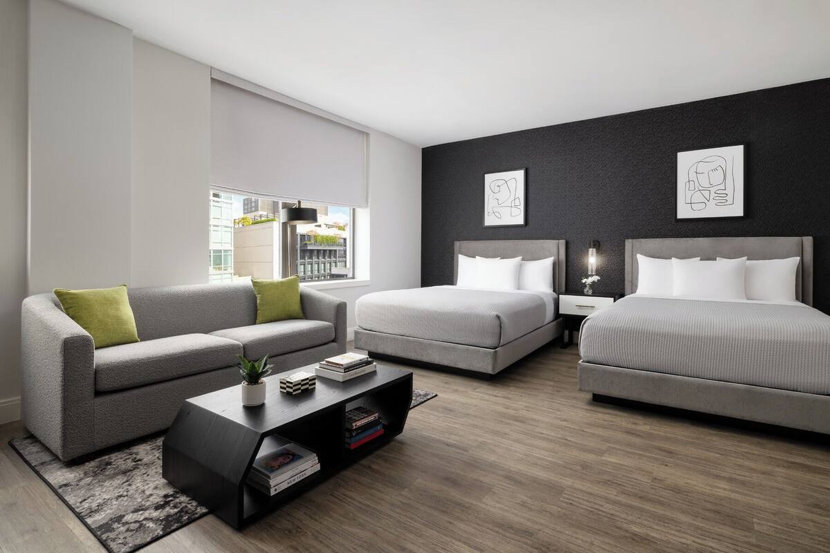 Quarto de hotel moderno com um sofá cinzento, duas camas de solteiro, uma mesinha de centro e vista para a cidade através de grandes janelas.