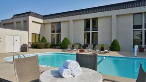 Área da piscina com espreguiçadeiras, uma mesa com toalhas e um edifício com janelas amplas e telhado plano.