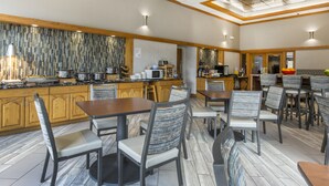 Uma sala de jantar ampla com mesas e cadeiras de madeira, área de cozinha com eletrodomésticos modernos e um papel de parede estampado.