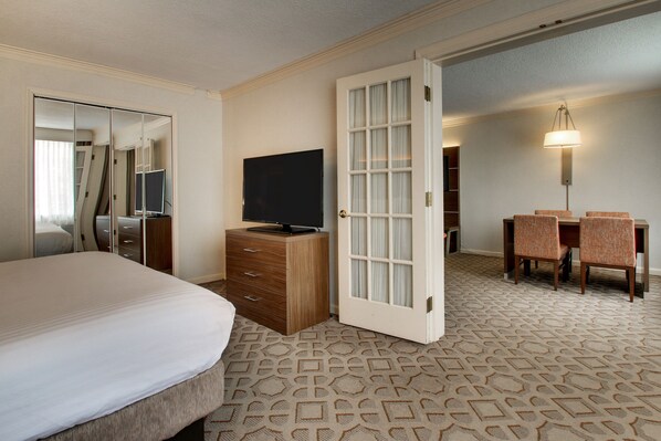 Quarto de hotel com cama, televisão sobre suporte de madeira, área de refeições com cadeiras e tapete estampado.