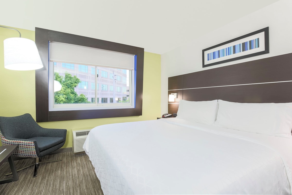Quarto de hotel com uma cama grande, uma cadeira, uma janela com vista para edifícios e uma moldura na parede.