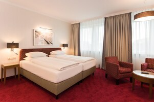 Quarto de hotel com duas camas, tapete vermelho, uma cadeira, uma mesinha e um quadro na parede.