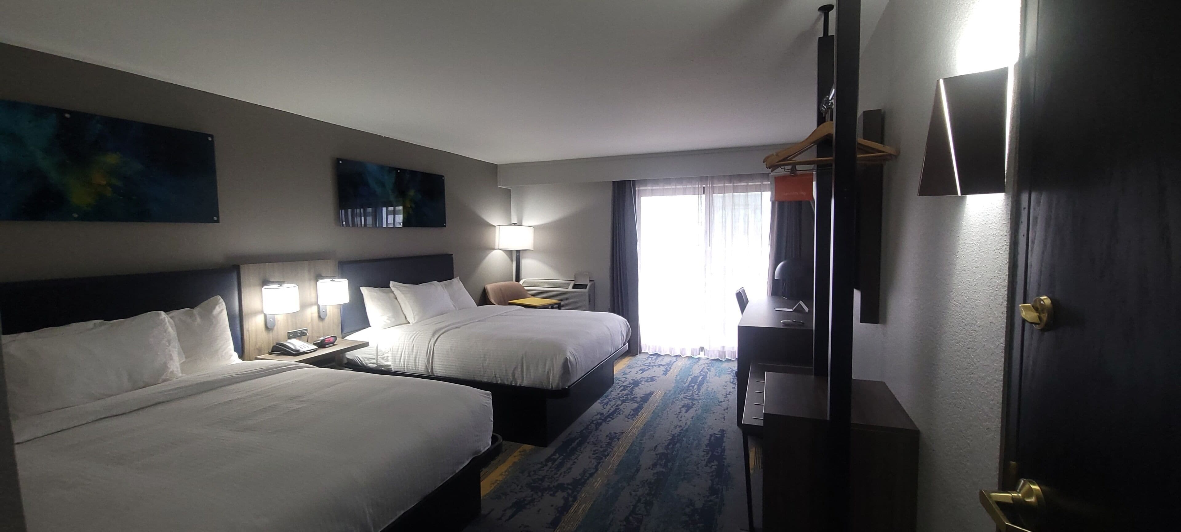 Quarto de hotel com duas camas, uma escrivaninha, uma luminária, televisão e uma janela grande com cortinas.