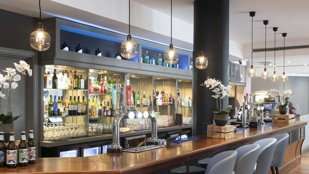 Uma área de bar com balcão de madeira, banquetas, luminárias pendentes e diversos tipos de garrafas e copos.