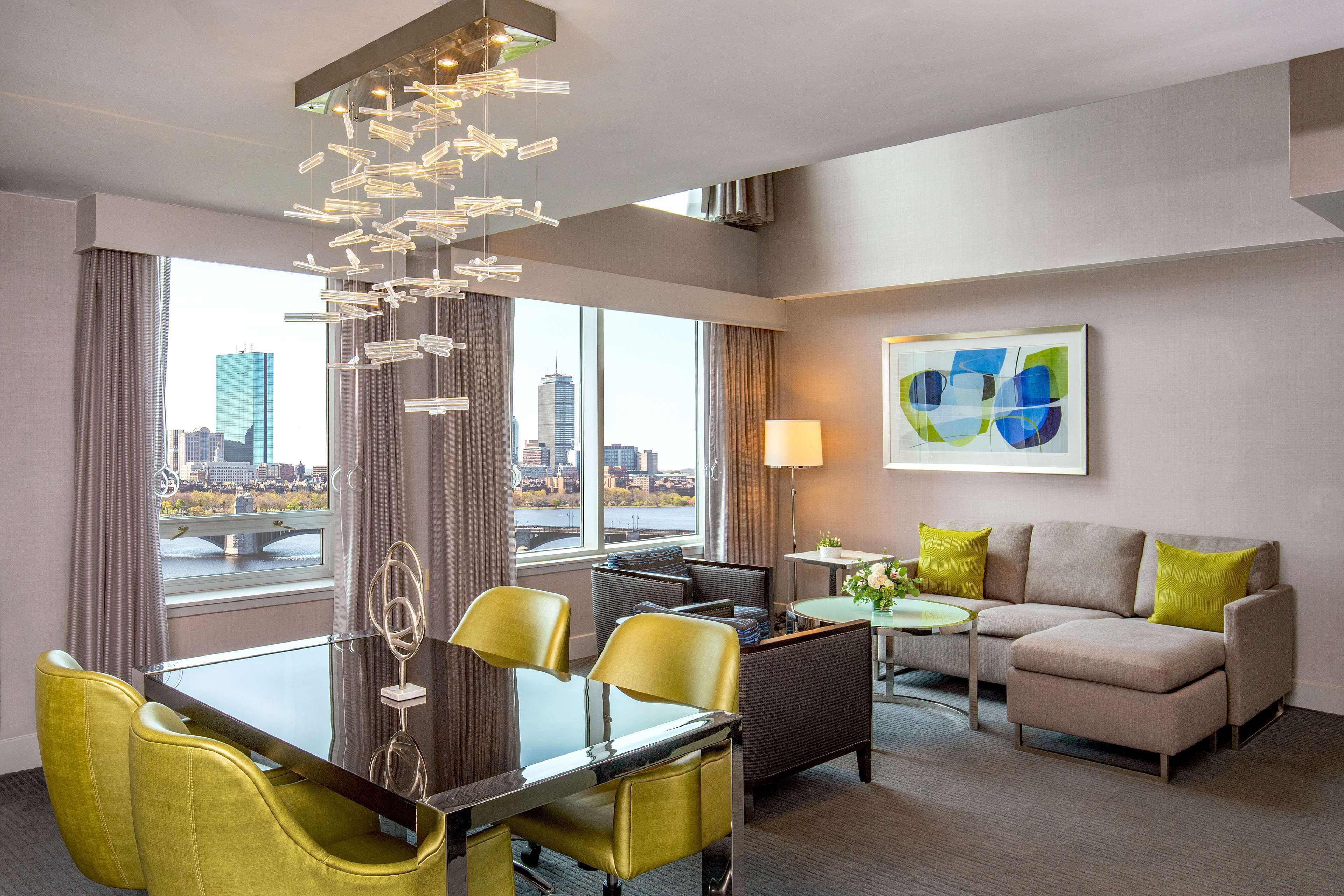 Quarto de hotel com área de jantar, sofá e uma grande janela com vista para a cidade.