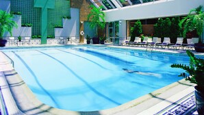 Uma piscina interior com teto de vidro, rodeada por espreguiçadeiras e plantas em vasos.