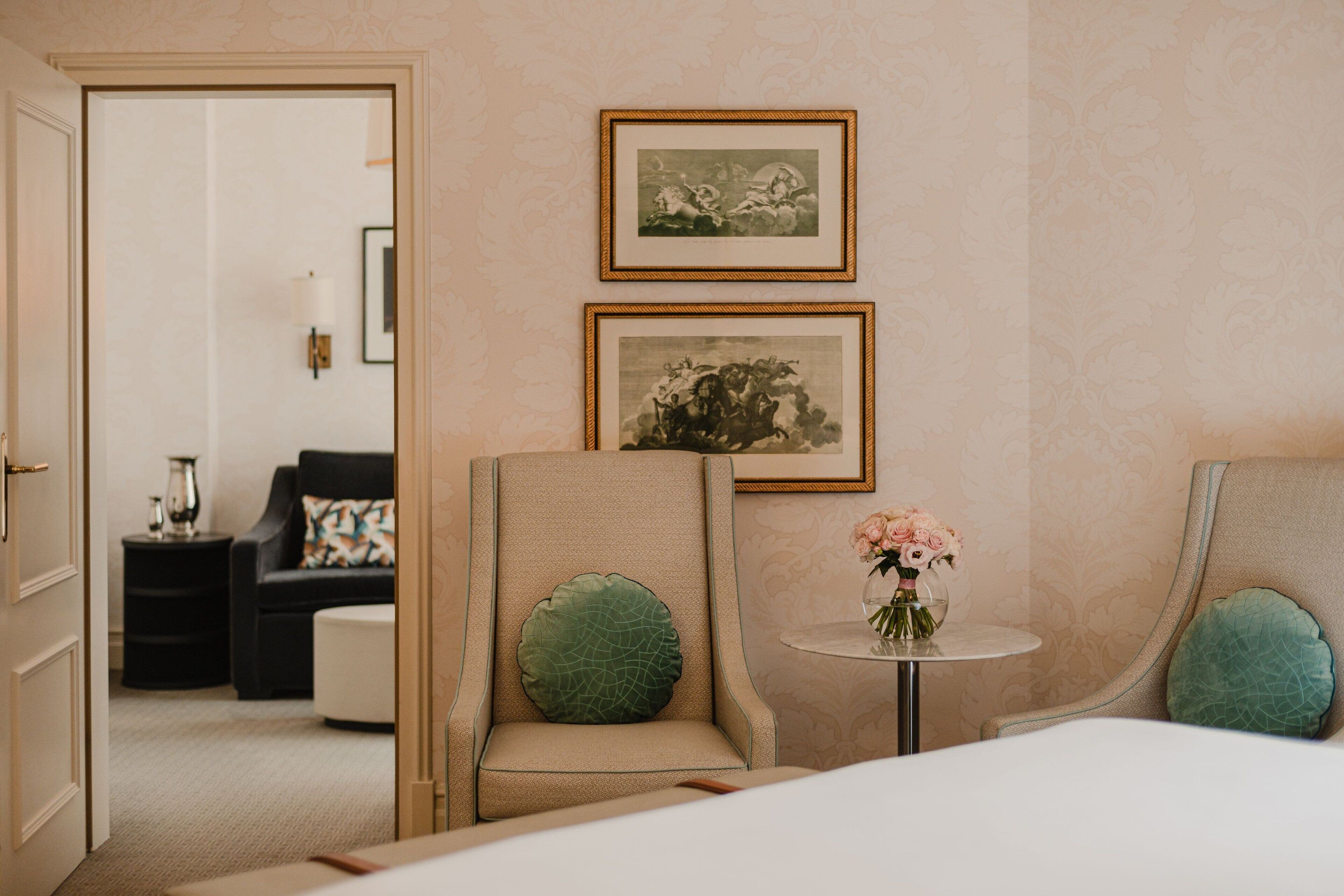 Quarto de hotel com cama, duas poltronas, uma mesinha de centro com um vaso de flores e duas fotos emolduradas na parede.