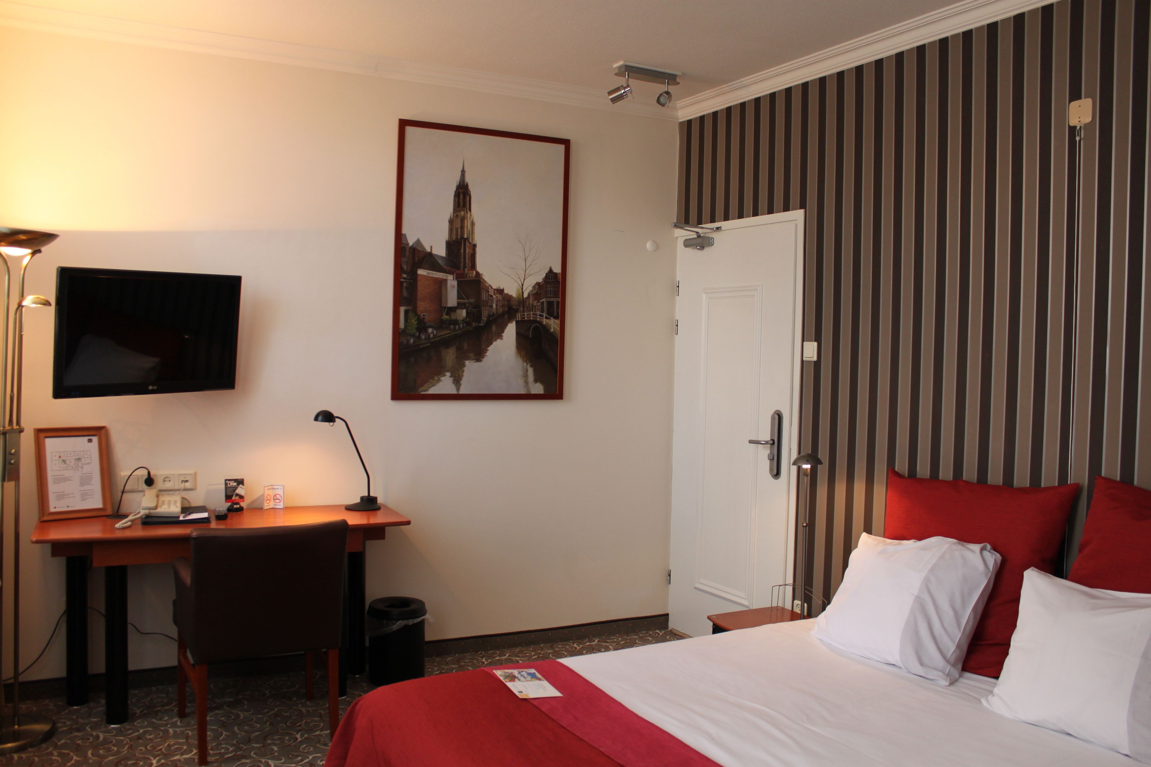 Quarto de hotel com cama, escrivaninha, cadeira, televisão presa na parede e quadro com vista para a cidade.