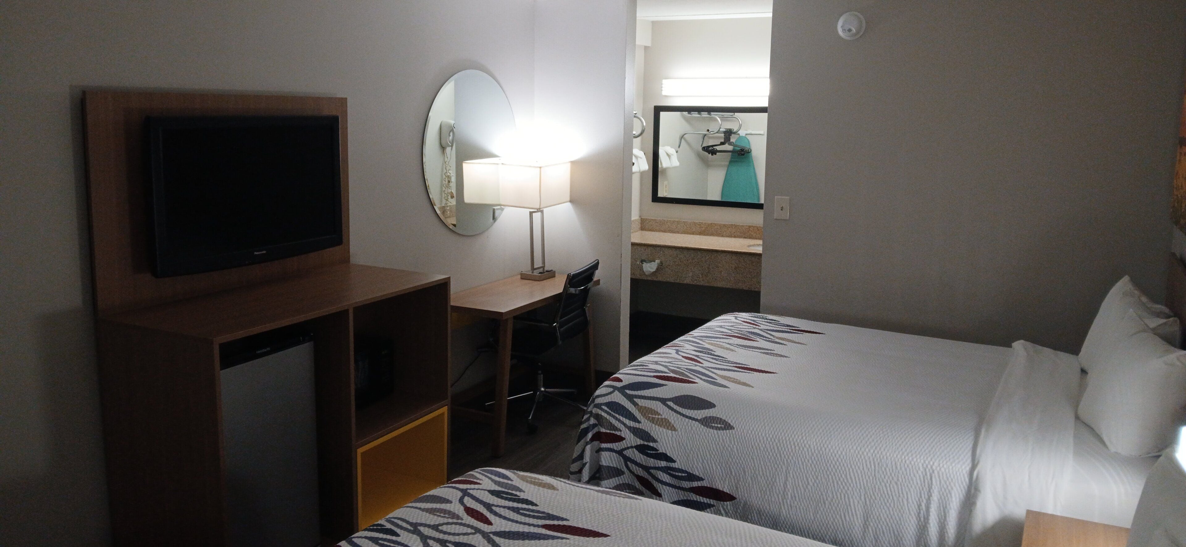 Quarto de hotel com duas camas, uma mesa com abajur, televisão e banheiro visível por uma porta aberta.