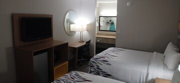 Quarto de hotel com duas camas, uma mesa com abajur, televisão e banheiro visível por uma porta aberta.