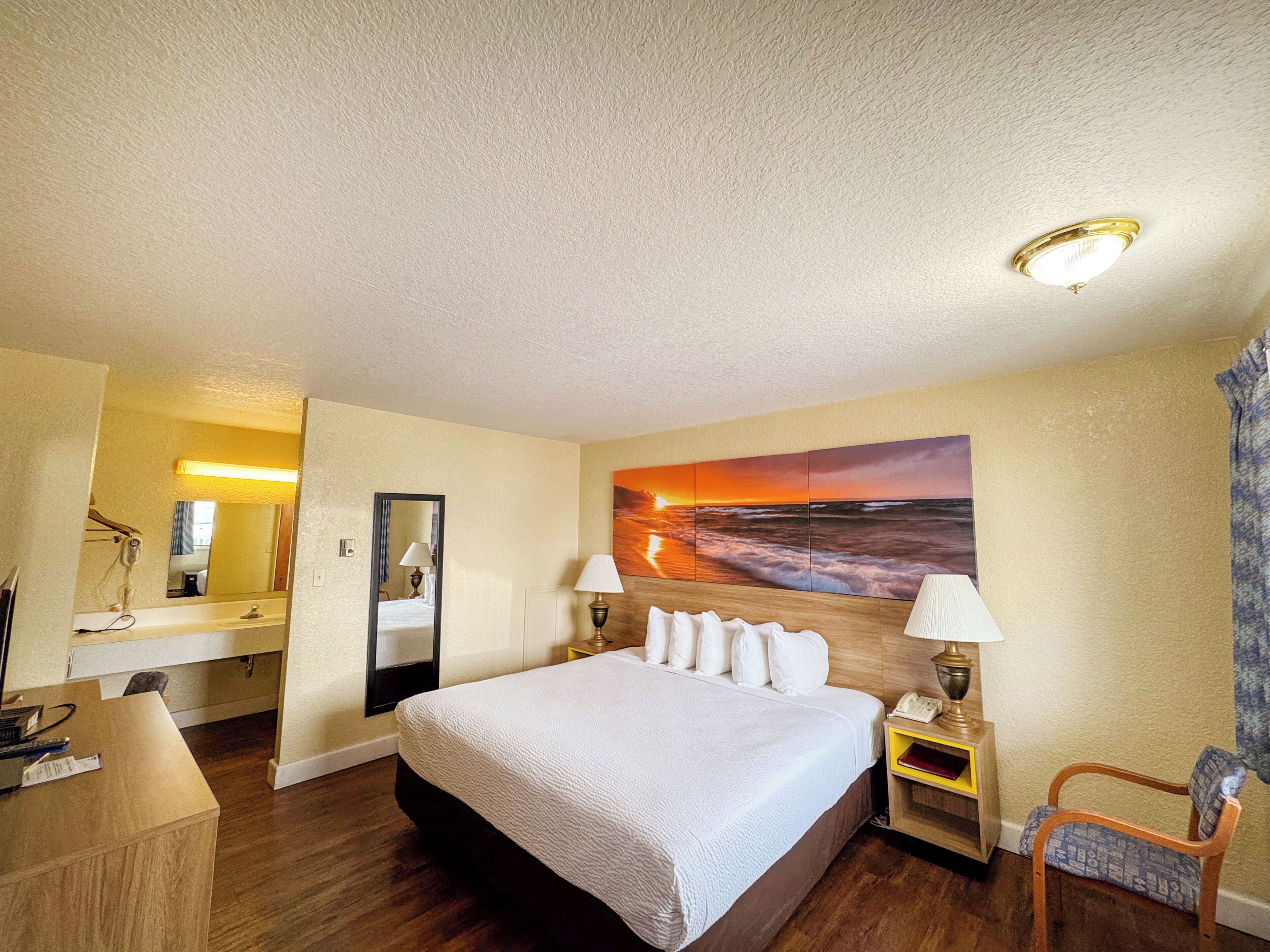 Quarto de hotel com uma cama grande, uma escrivaninha, uma cadeira, um quadro de um pôr do sol e uma luminária no teto.