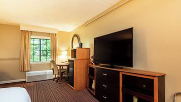 Quarto de hotel com cama, escrivaninha, televisão, cadeira, luminária e janela com cortinas.