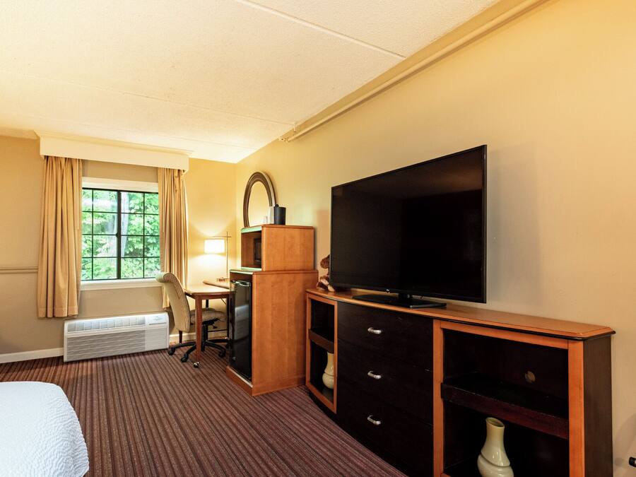 Quarto de hotel com cama, escrivaninha, televisão, cadeira, luminária e janela com cortinas.