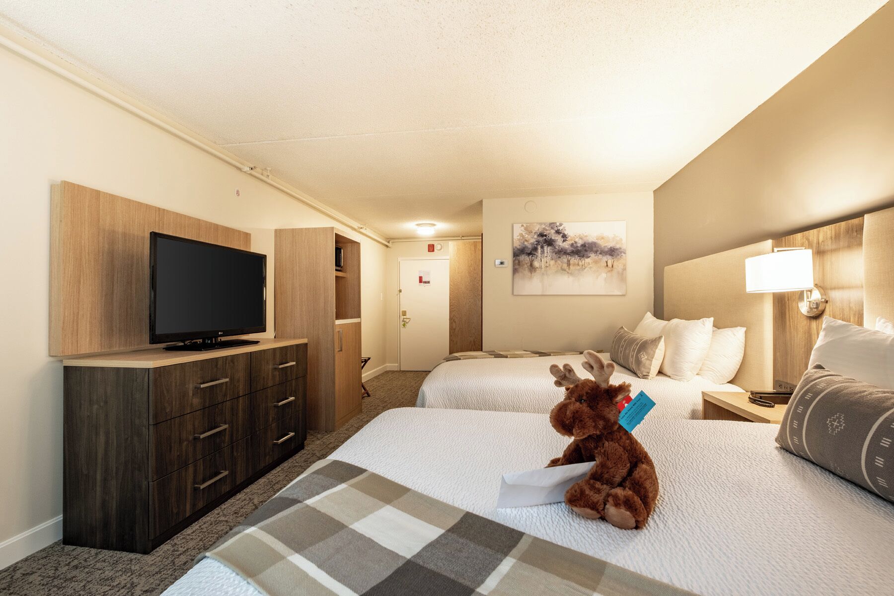 Quarto de hotel com televisão de tela plana, guarda-roupa de madeira, duas camas com roupa de cama branca, um quadro na parede e uma luminária na mesa de cabeceira.