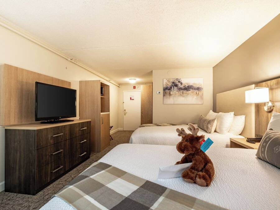 Quarto de hotel com televisão de tela plana, guarda-roupa de madeira, duas camas com roupa de cama branca, um quadro na parede e uma luminária na mesa de cabeceira.