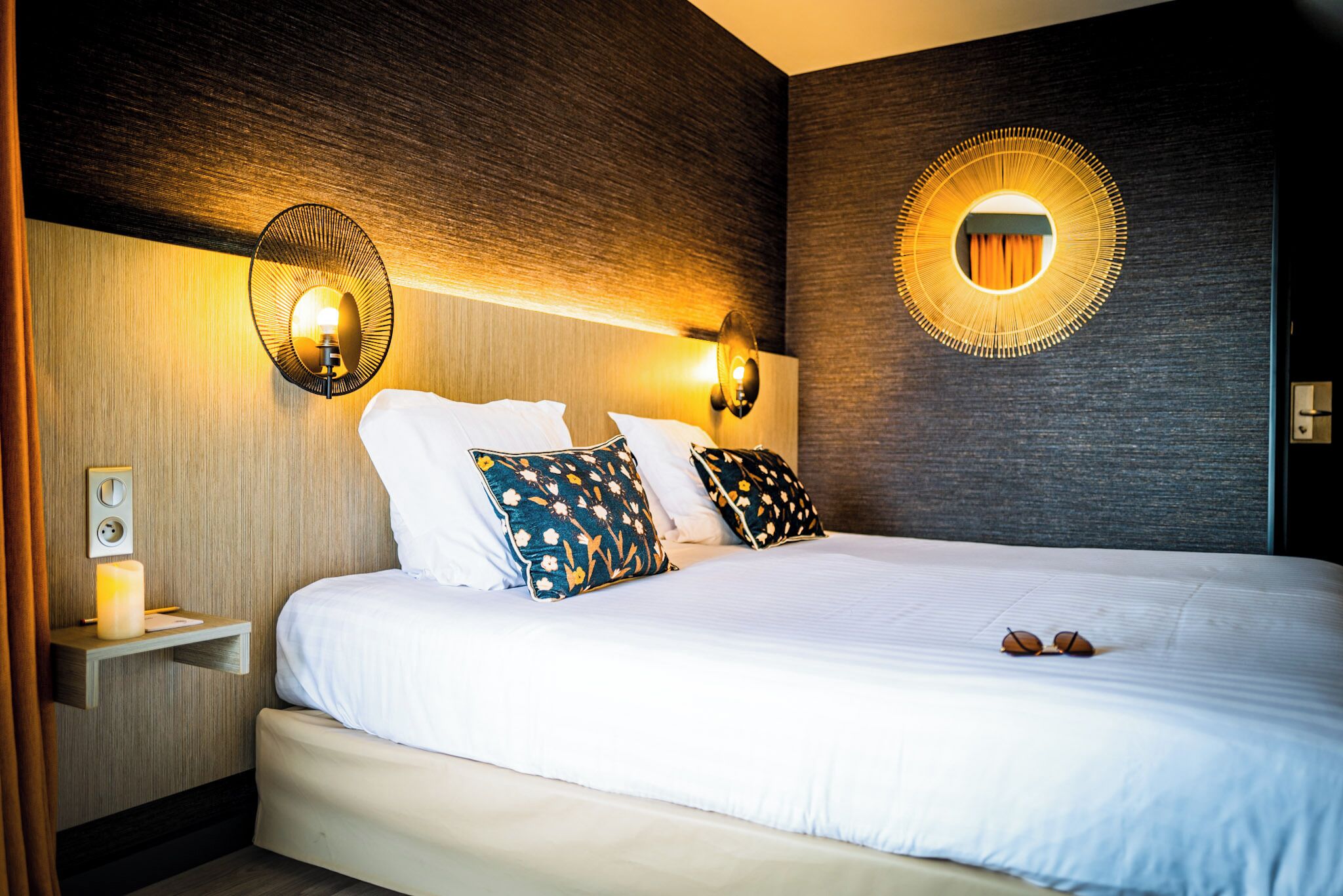 Quarto de hotel moderno com uma cama grande, duas arandelas, uma mesa de cabeceira com abajur e um espelho circular na parede.