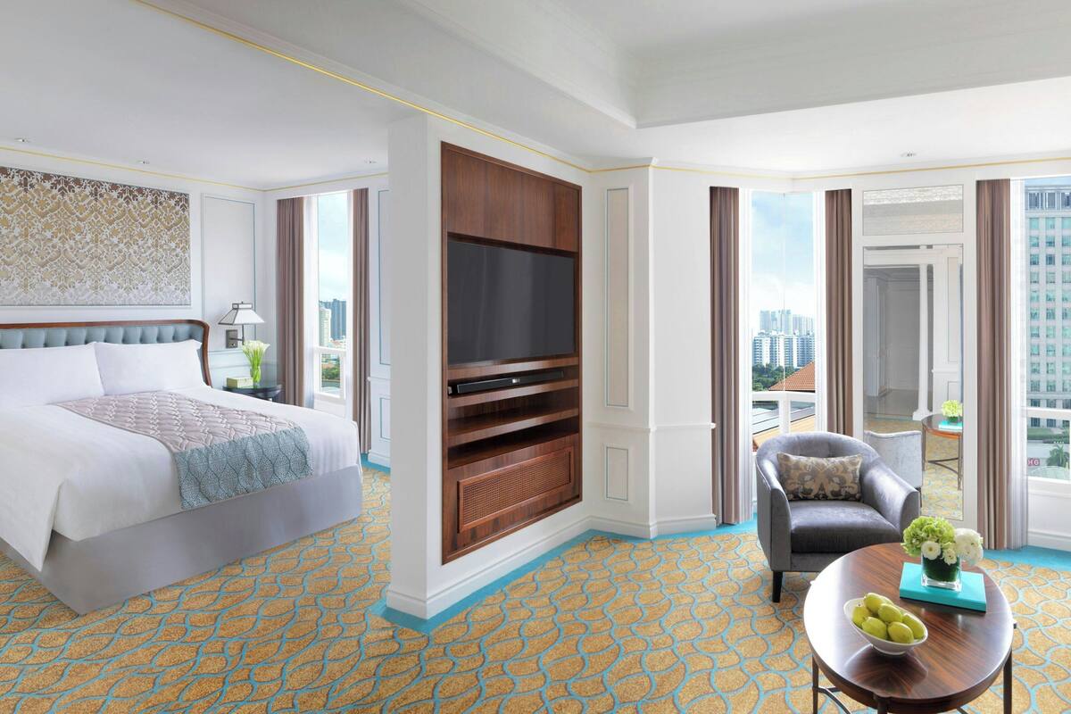 Quarto de hotel com cama, televisão, poltrona, mesa com um vaso e carpete estampado.