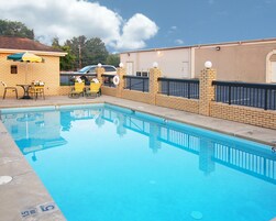 Piscina com muro de tijolos e cerca, área coberta com cadeiras, e um edifício ao fundo.
