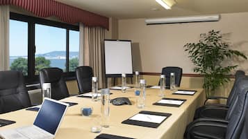 Sala de conferência com uma mesa comprida, cadeiras pretas, um laptop, garrafas de água e um quadro branco.