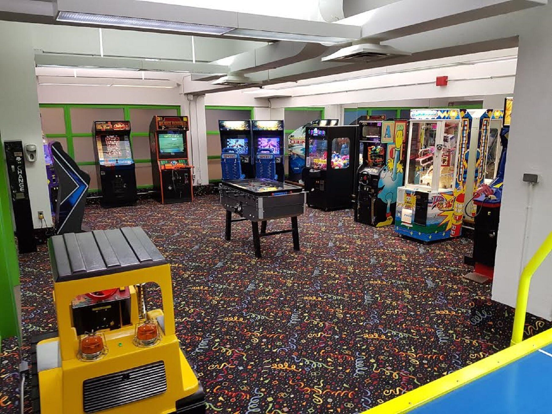 Um fliperama com diversas máquinas de jogos, uma máquina de vendas amarela e piso coberto de carpete.