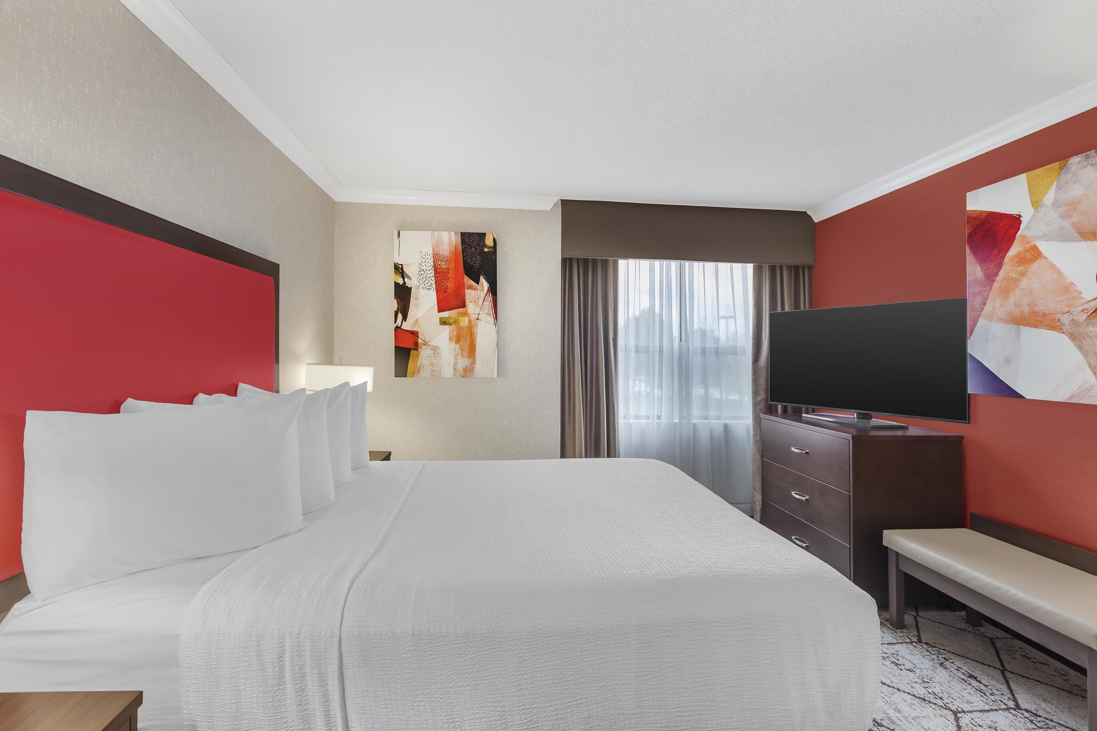 Quarto de hotel com cama grande, televisão, um banquinho e um quadro na parede.