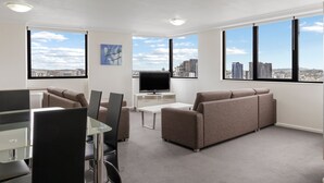 Uma sala de estar moderna com uma mesa de jantar de vidro, dois sofás, uma televisão e grandes janelas com vista para a cidade.