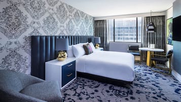 Quarto de hotel com cama grande, mesa de cabeceira com luminárias, uma cadeira, uma pequena mesa com uma cadeira e vista da cidade pela janela.