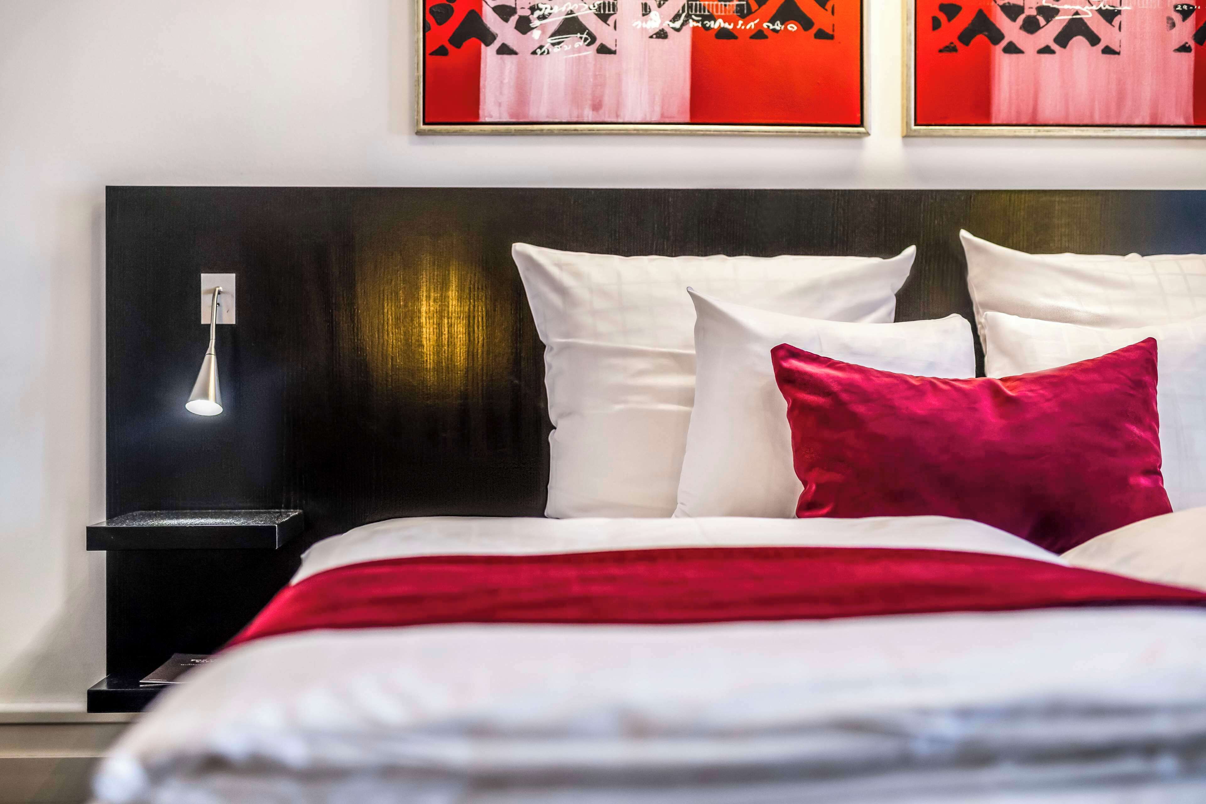 Uma cama bem arrumada com roupa de cama branca e vermelha, um criado-mudo preto com uma luminária, e uma parede com arte abstrata.