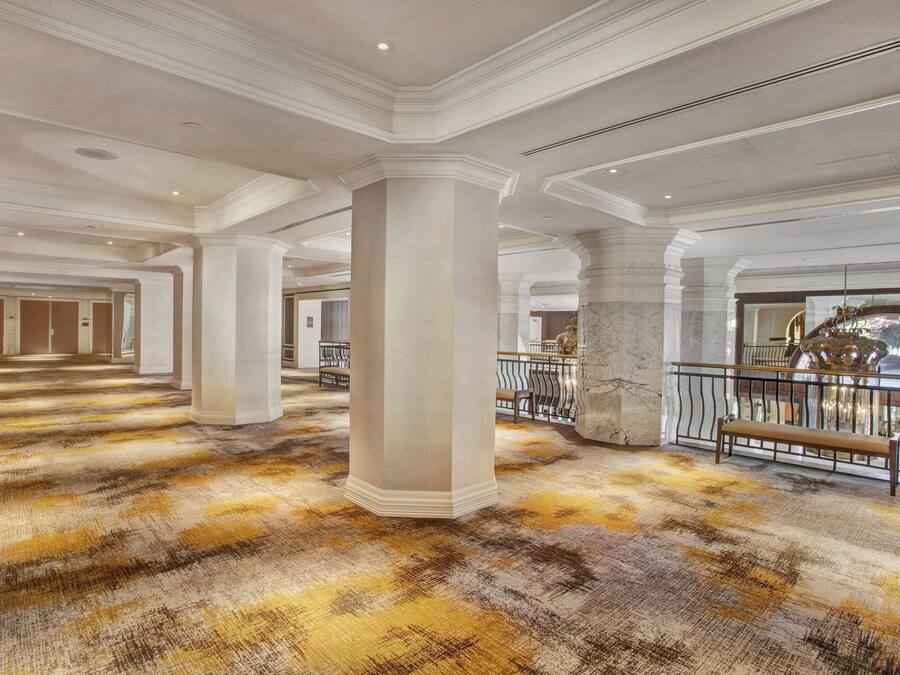 Um hall de hotel espaçoso, com pé direito alto, colunas de mármore e um tapete estampado.