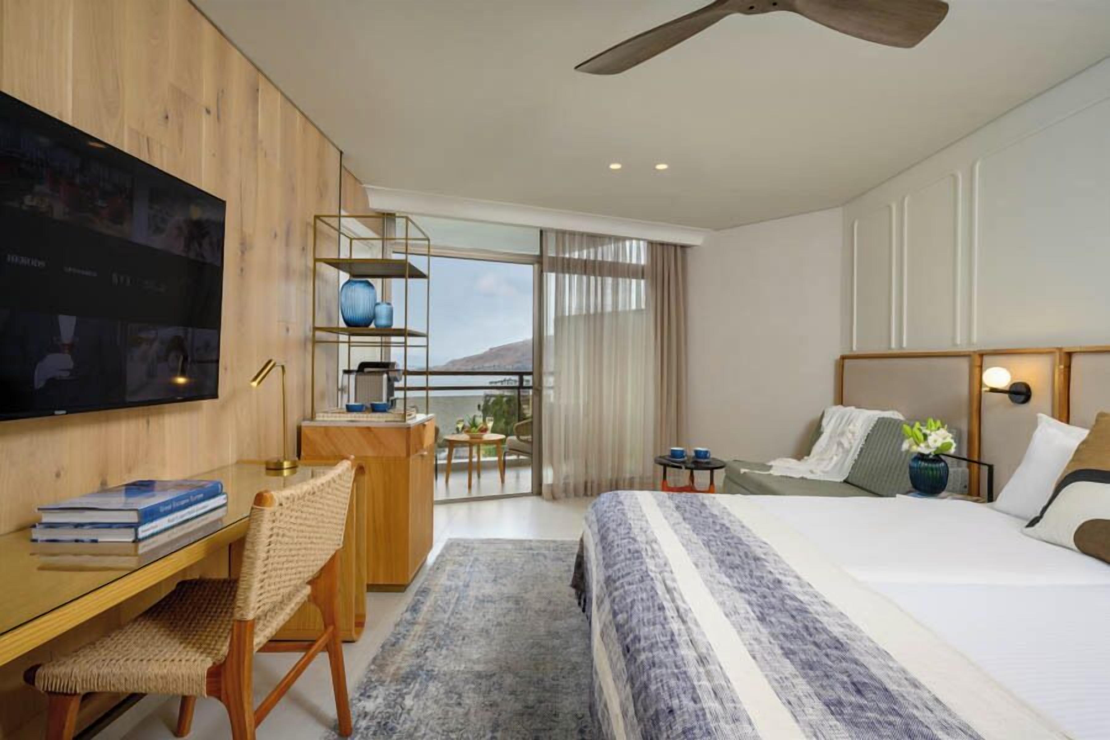 Quarto de hotel moderno com uma cama grande, uma escrivaninha com cadeira, TV e varanda com vista.