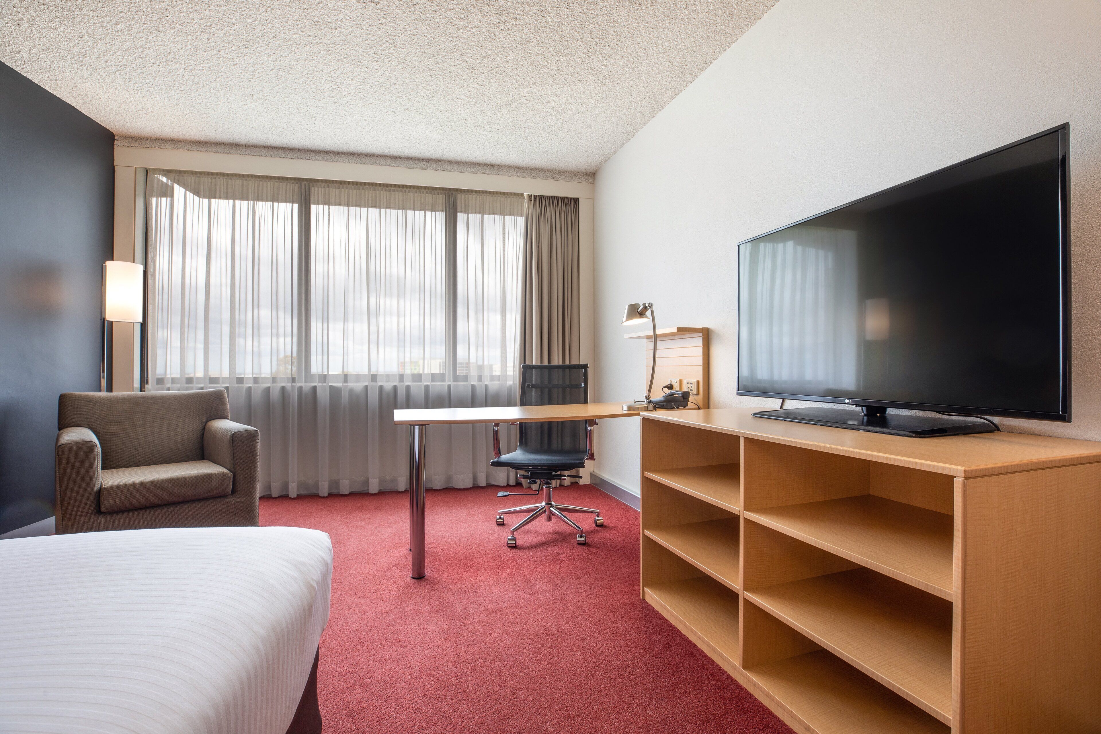 Quarto de hotel com cama, escrivaninha, cadeira, televisão e uma janela grande com cortinas.