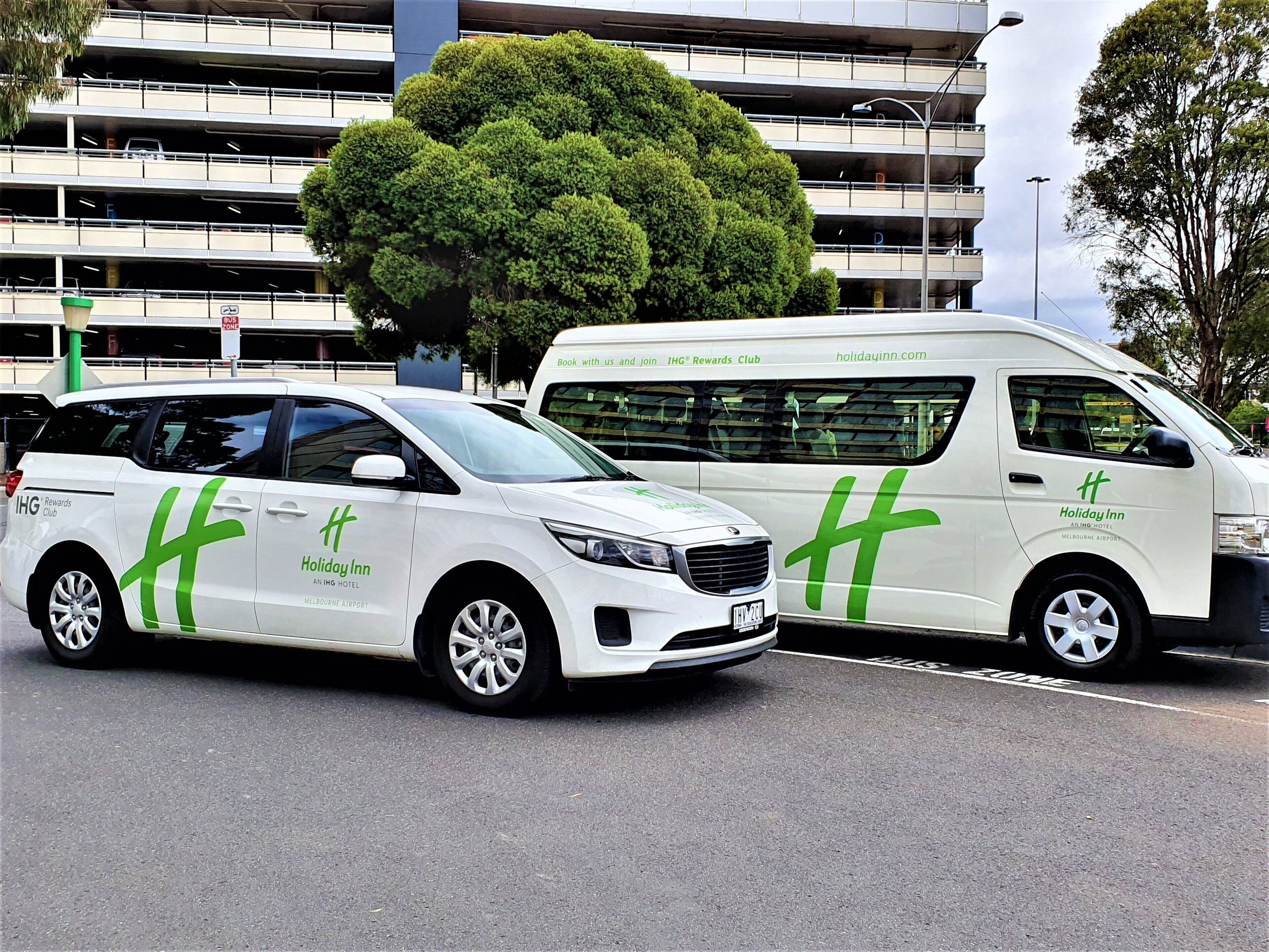 Uma carrinha branca e um carro branco com letras verdes, estacionados em frente a um edifício com vários níveis de estacionamento e uma grande árvore.