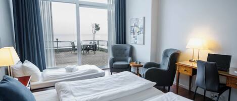 Quarto de hotel com duas camas, uma escrivaninha, duas cadeiras, um laptop, uma luminária e vista para o mar através de amplas janelas.